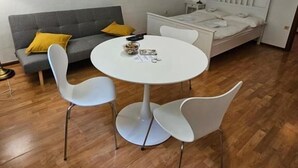 Quarto pequeno e moderno com sofá cinzento, mesa redonda branca com duas cadeiras, cama com cabeceira branca e luminária suspensa.