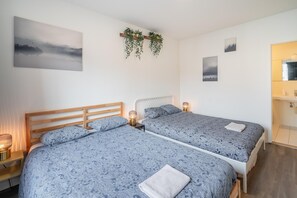 Quarto com duas camas, mesa de cabeceira com abajur, prateleira na parede com plantas e um quadro.
