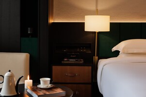 Quarto de hotel com uma cama, uma mesa de cabeceira com abajur, uma chávena, um livro e uma vela.