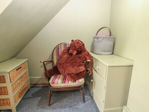 Um quarto com teto inclinado, uma cadeira com almofada listrada, um urso de pelúcia grande, uma cesta de vime e um armário branco.