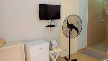 Quarto com televisão na parede, um ventilador, um pequeno frigorífico branco e uma porta que dá para um espaço de duche.