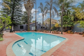 Piscina com borda azulejada, rodeada por um terraço de tijolos, palmeiras e mobiliário de exterior.