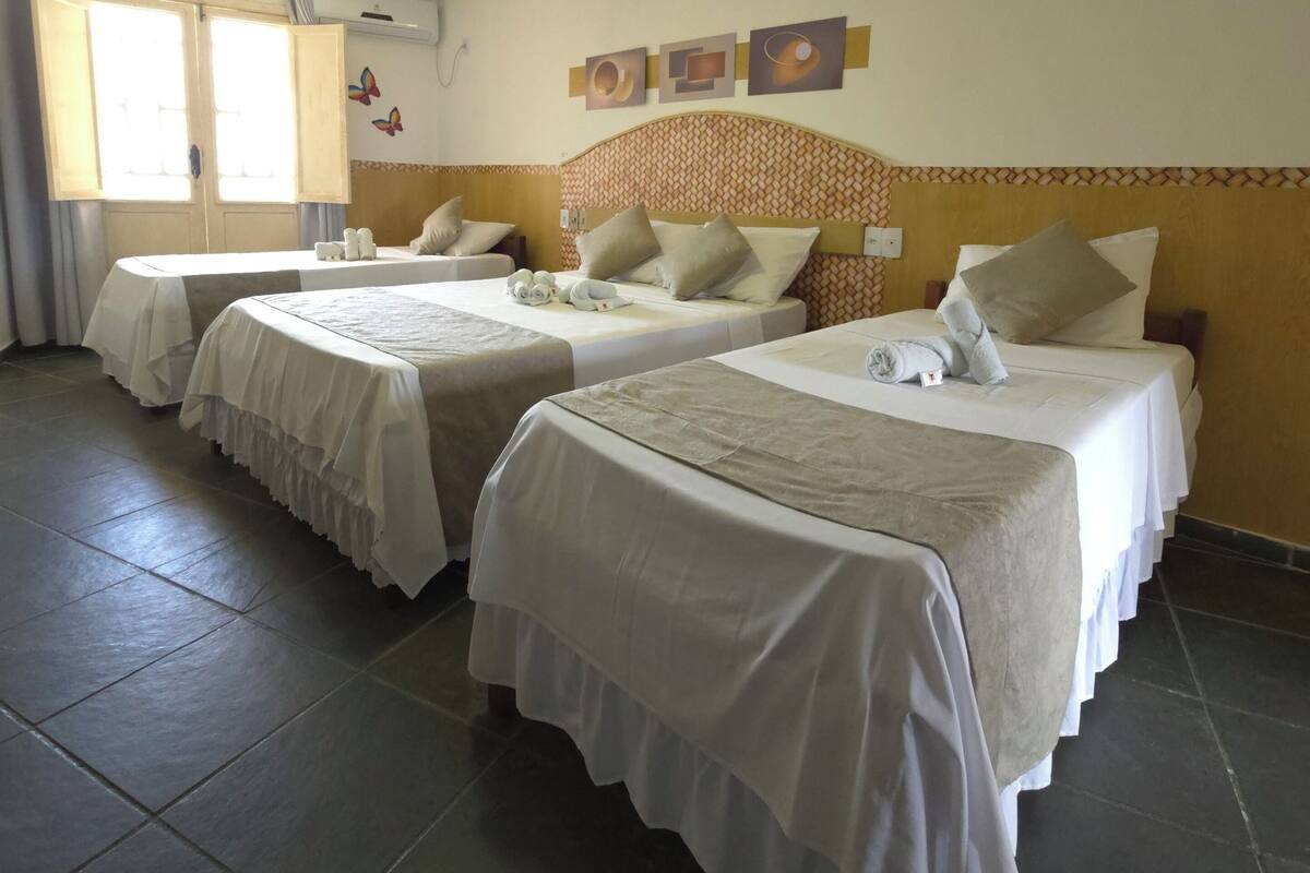 Quarto de hotel com três camas, janela com cortinas, ar-condicionado fixado na parede e luminária no teto.