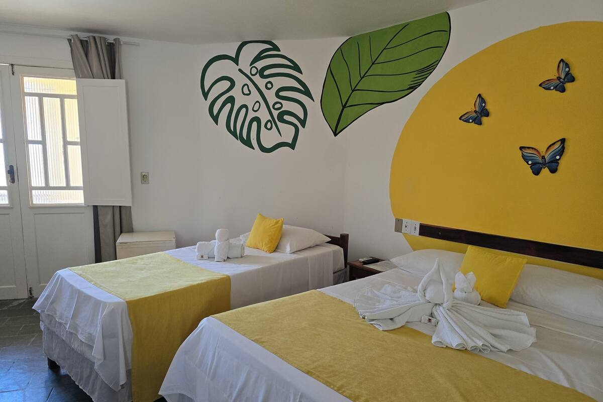 Duas camas de solteiro com roupa de cama branca e amarela, uma parede com decoração de folhas e borboletas, e uma janela com cortinas.
