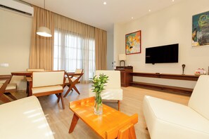 Quarto de hotel com sofá, mesa de centro de madeira com um vaso, mesa de jantar com cadeiras, televisão de tela plana e um quadro na parede.