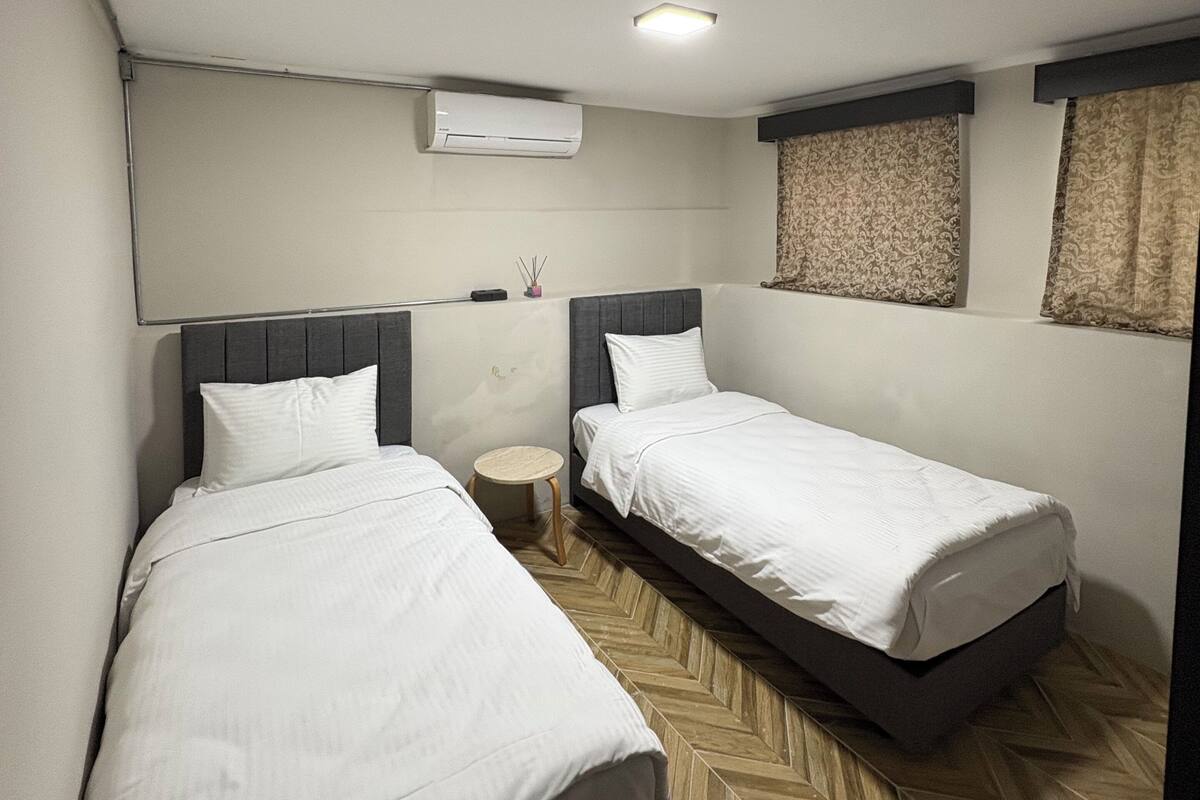 Quarto de hotel com duas camas, uma mesa redonda pequena, um aparelho de ar condicionado e uma luminária de teto.