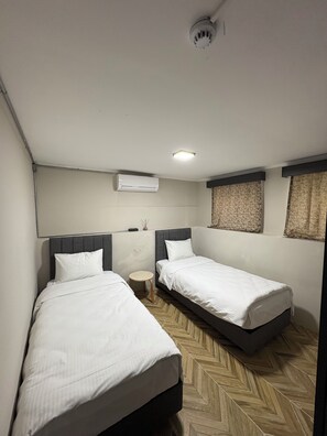 Quarto de hotel com duas camas, uma mesa redonda pequena, um aparelho de ar condicionado e uma luminária de teto.