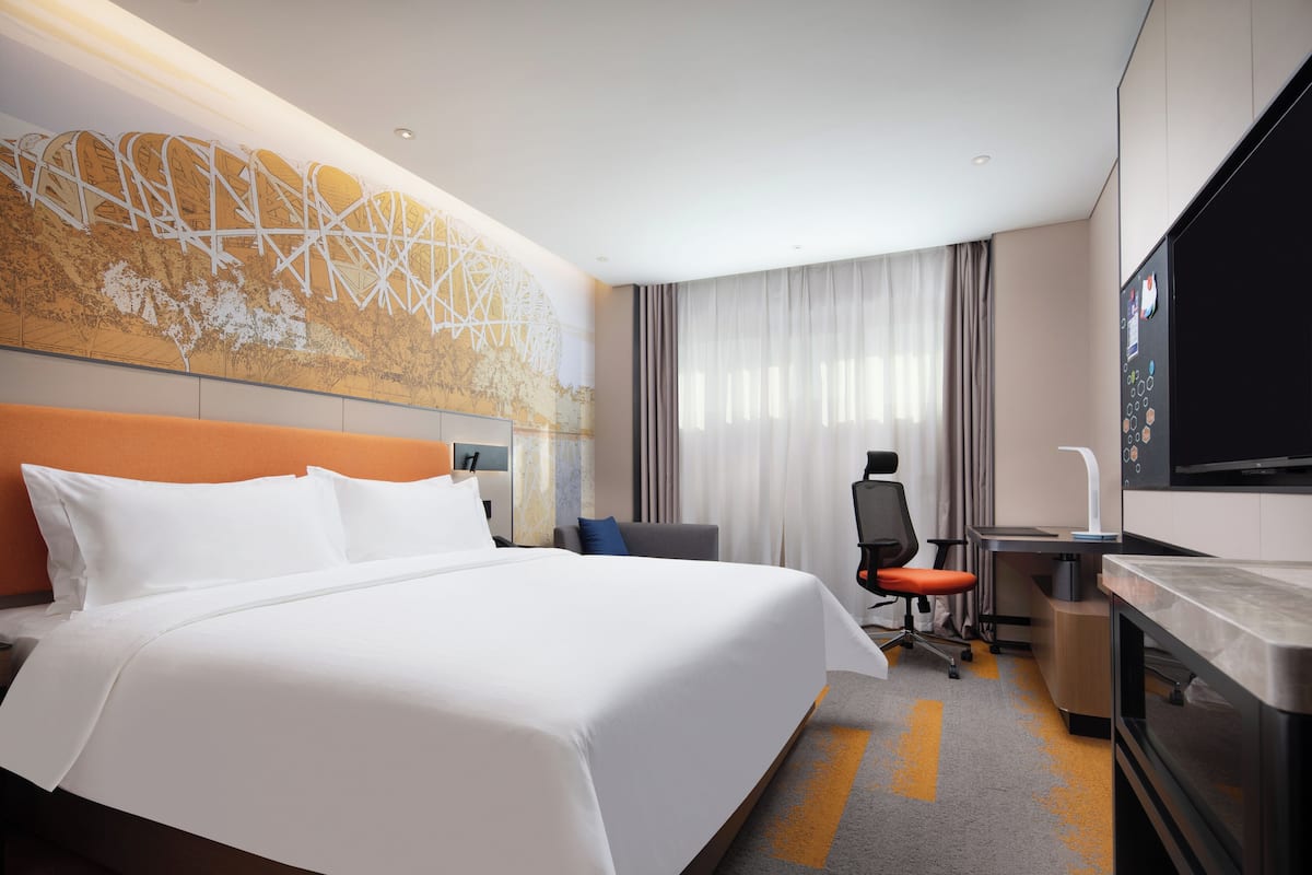 Quarto de hotel com uma cama grande, uma escrivaninha, uma cadeira, uma televisão e um detalhe decorativo na parede.