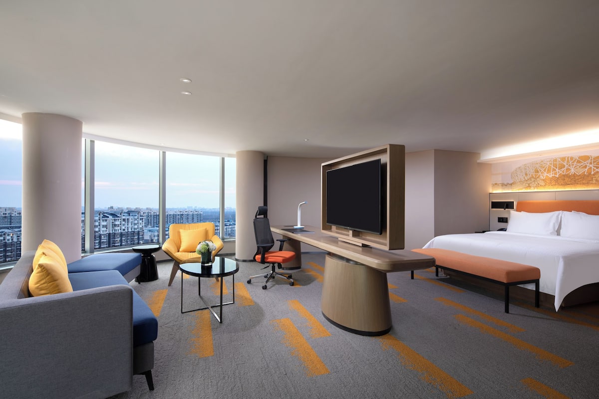 Quarto de hotel moderno com uma cama grande, uma escrivaninha, uma cadeira, um sofá e uma televisão.