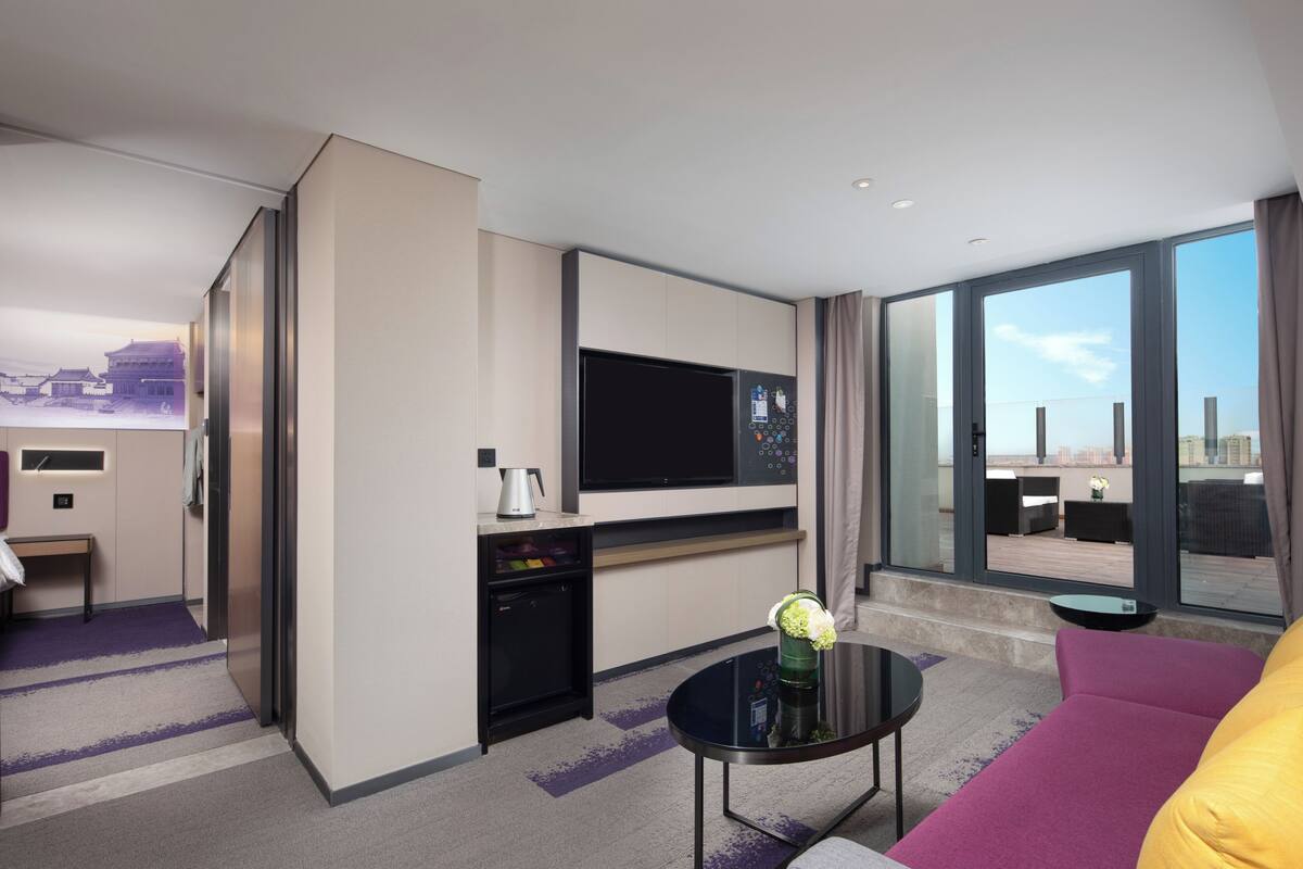 Quarto de hotel moderno com uma televisão grande, uma pequena cozinha compacta, um sofá e uma varanda com vista para a cidade.
