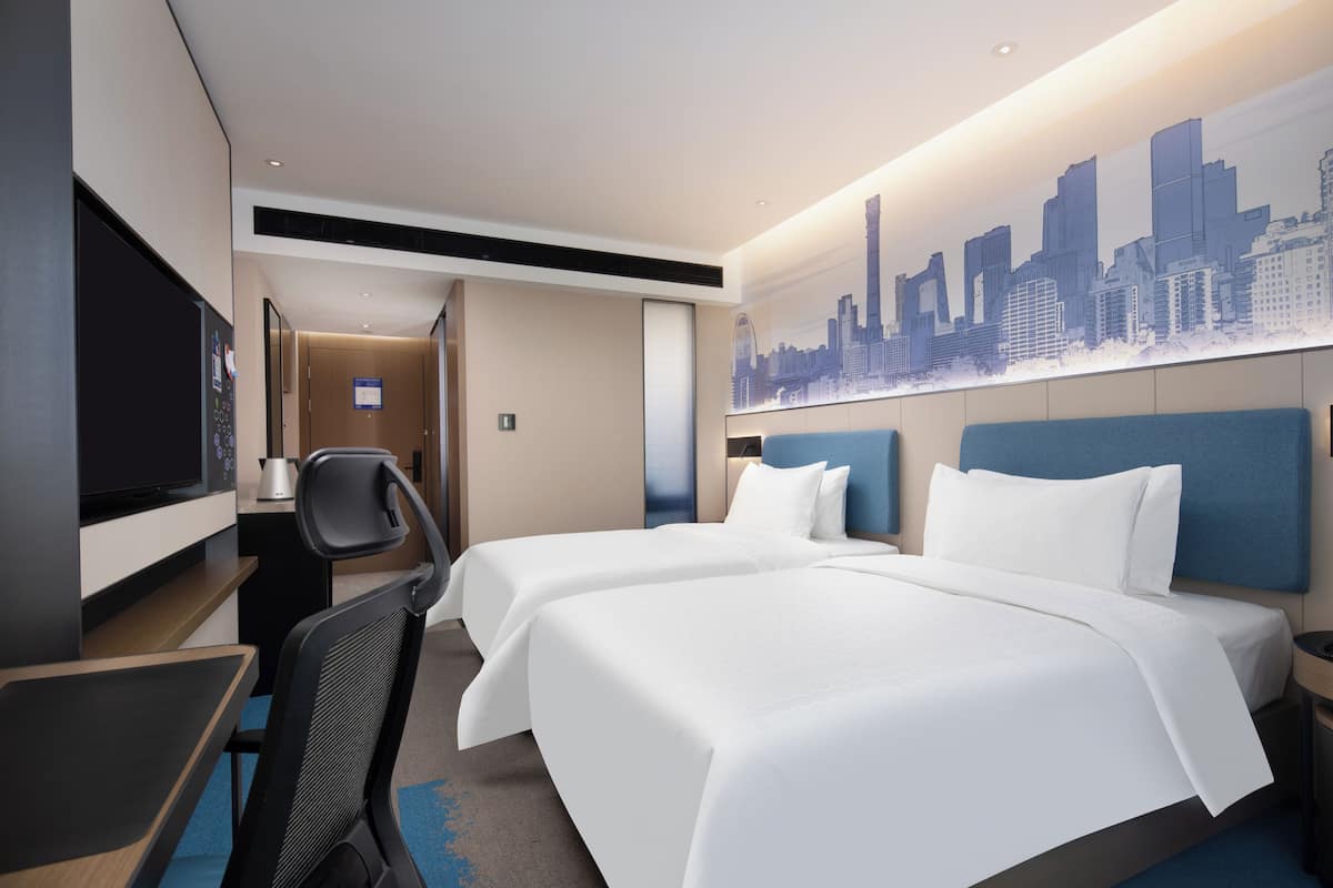 Quarto de hotel com duas camas, uma escrivaninha, uma cadeira, uma televisão e um mural com vista para a cidade.