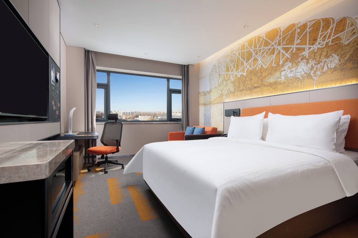 Quarto de hotel com cama, escrivaninha com cadeira, televisão e uma grande janela com vista para a cidade.
