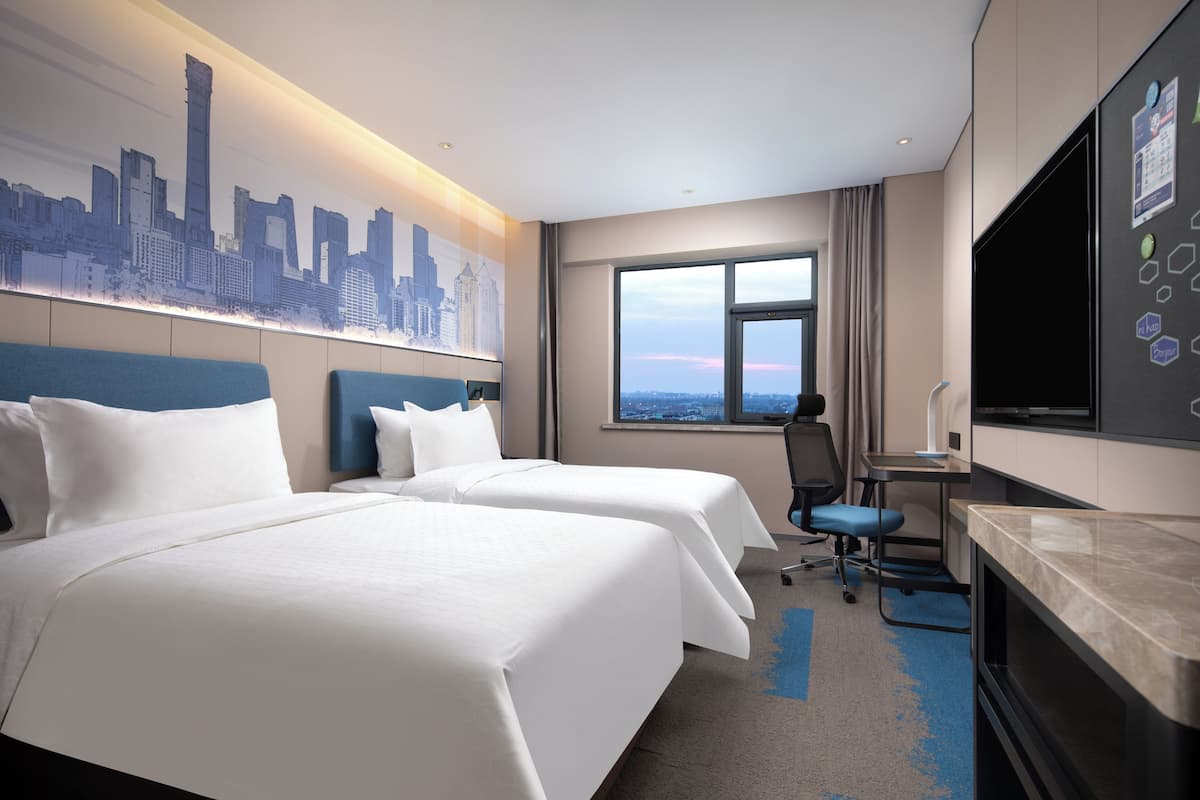 Quarto de hotel com duas camas, uma escrivaninha, uma cadeira, uma TV, uma janela com vista para a cidade e um mural na parede retratando o horizonte urbano.