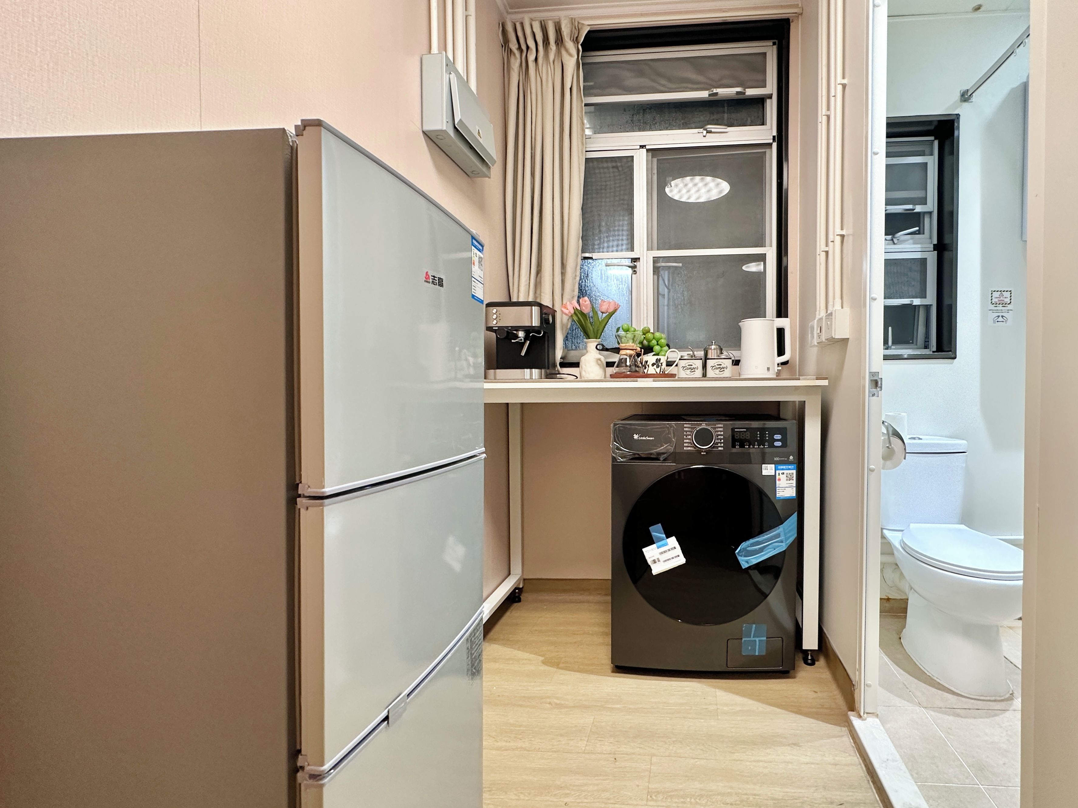 Cozinha compacta com geladeira, máquina de lavar e janela com cortinas.