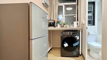 Cozinha compacta com geladeira, máquina de lavar e janela com cortinas.