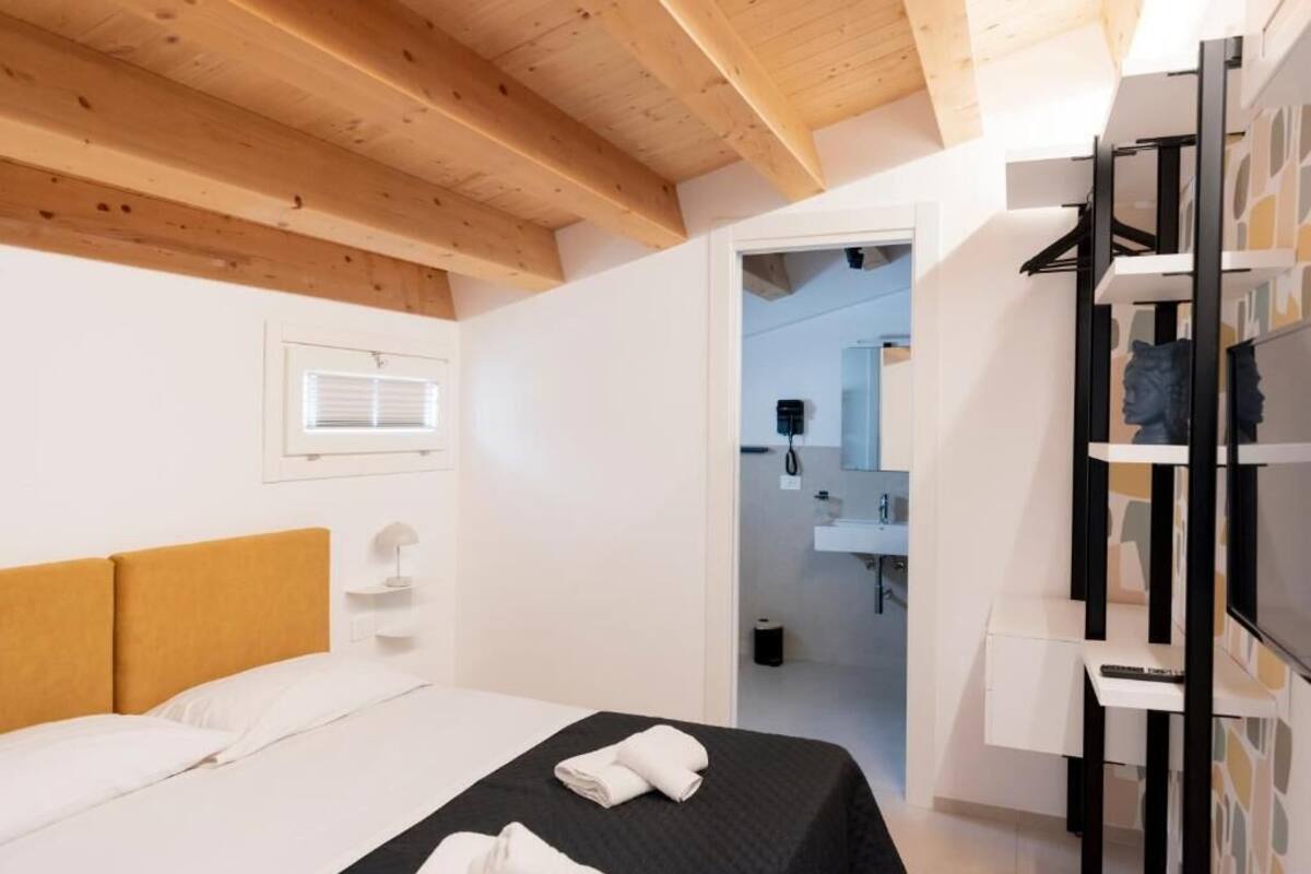 Quarto com cama, teto de madeira, parede branca, porta para um banheiro e uma estante preta.