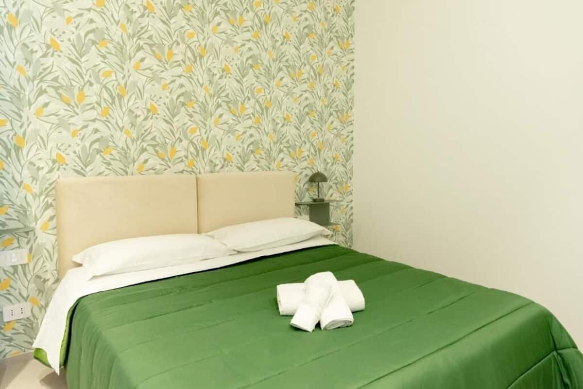 Uma cama com colcha verde e almofadas brancas, uma pequena mesa de cabeceira com abajur, e um papel de parede com padrão floral.