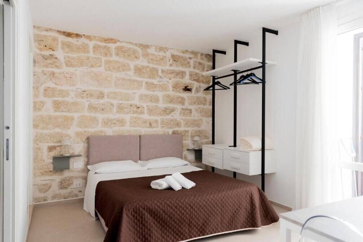 Um quarto com parede de pedra, uma cama com colcha marrom, uma mesinha de cabeceira, um cabideiro e um aparador branco.