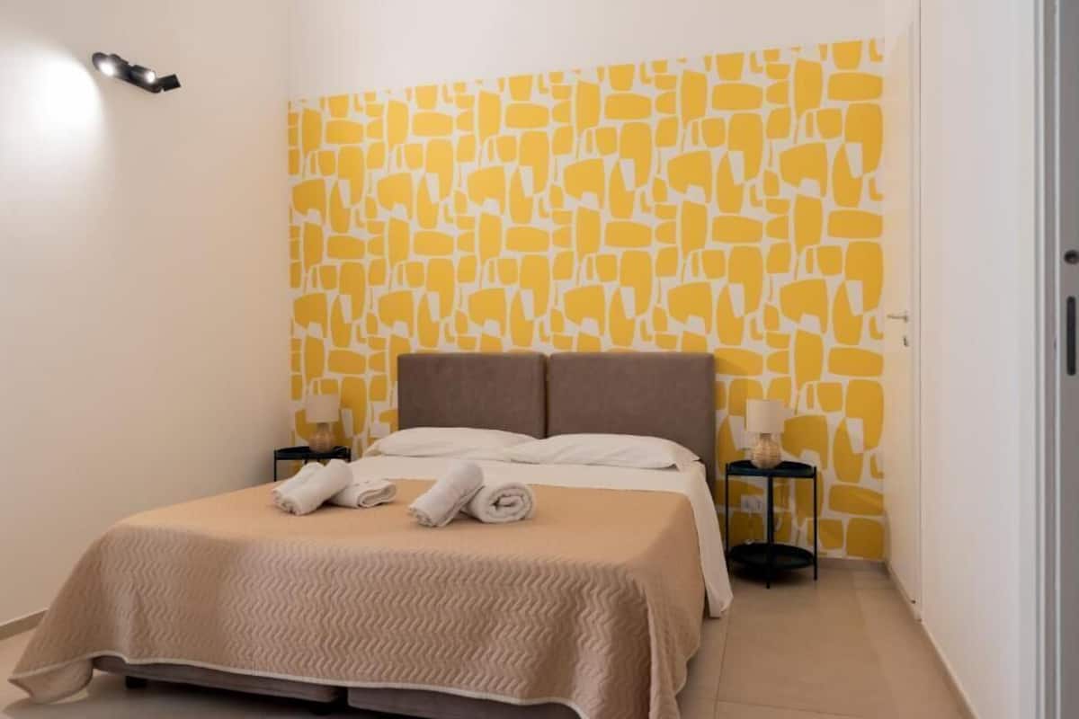 Quarto de hotel com uma cama, duas mesas de cabeceira, uma parede com padrão abstrato amarelo e uma porta.