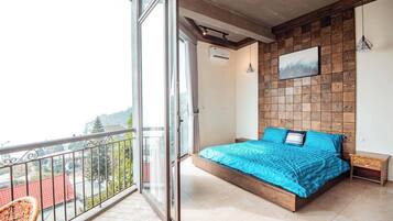 Quarto com cama grande, cabeceira de madeira, varanda com cadeira e vista para a cidade.