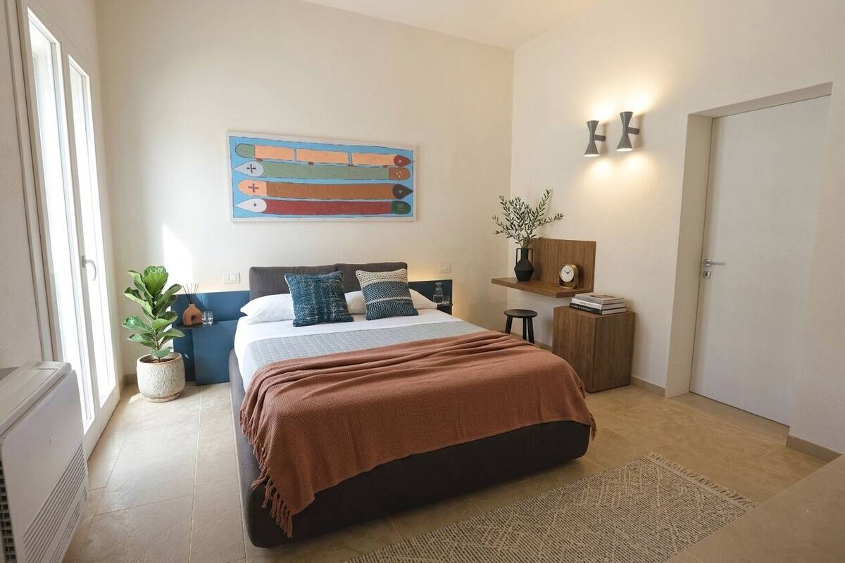 Um quarto com uma cama, mesas de cabeceira, um relógio na parede, uma planta em vaso e um quadro.
