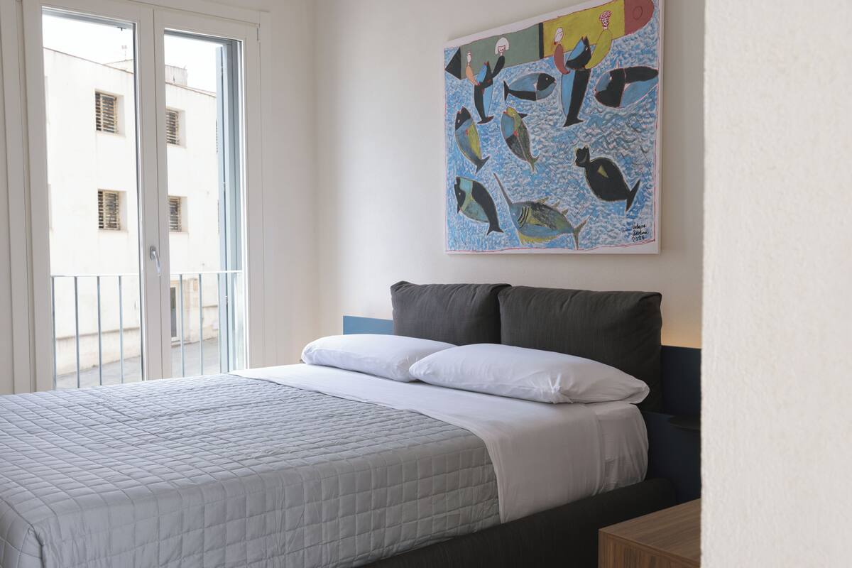 Quarto com cama, mesa de cabeceira, um quadro e vista pela janela.
