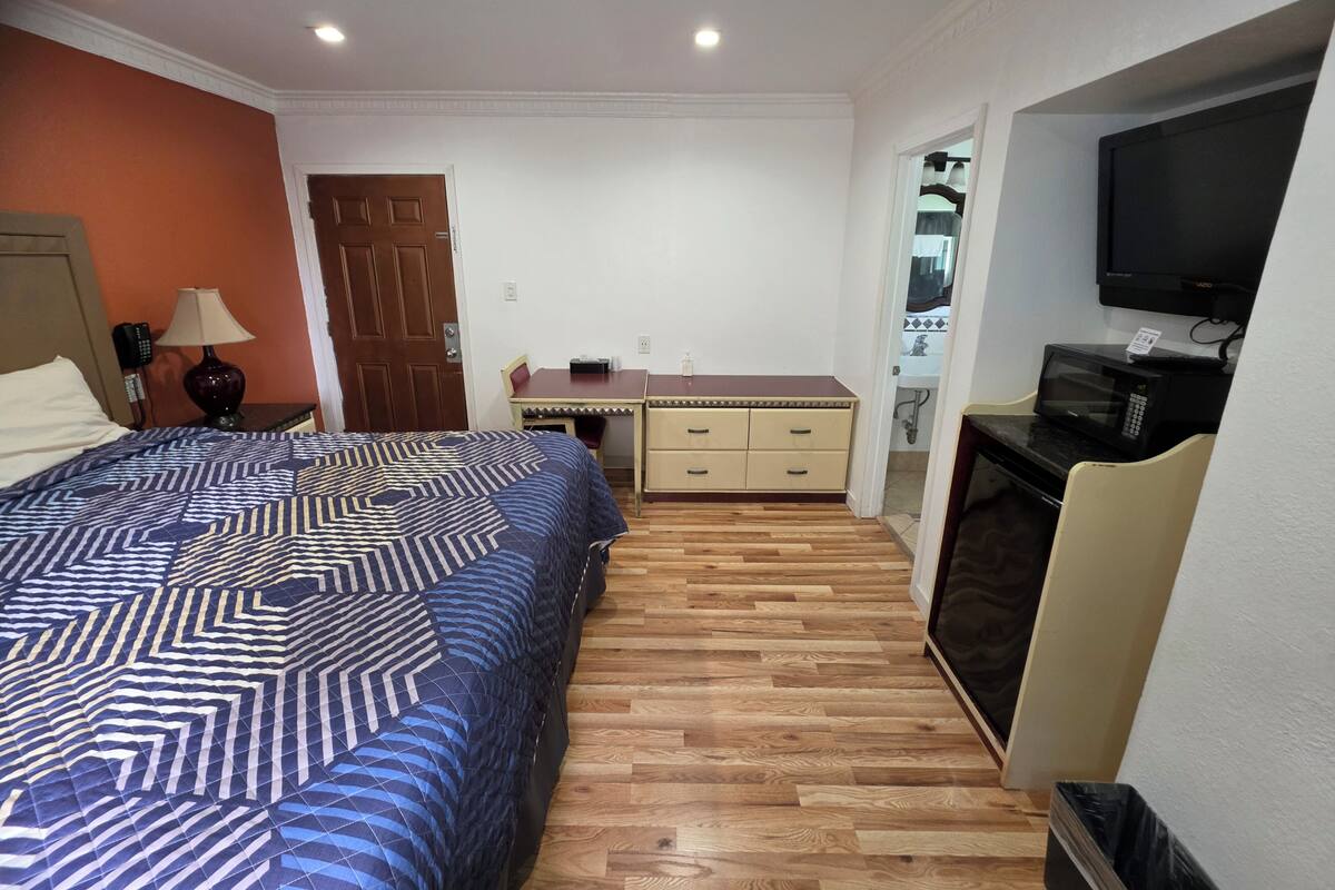Quarto de hotel com cama, escrivaninha, cadeira, televisão e banheiro visível por uma porta aberta.