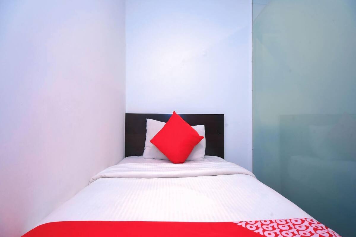 Uma cama de solteiro com colchão branco e travesseiro vermelho, cabeceira preta e edredom com padrão vermelho e branco.
