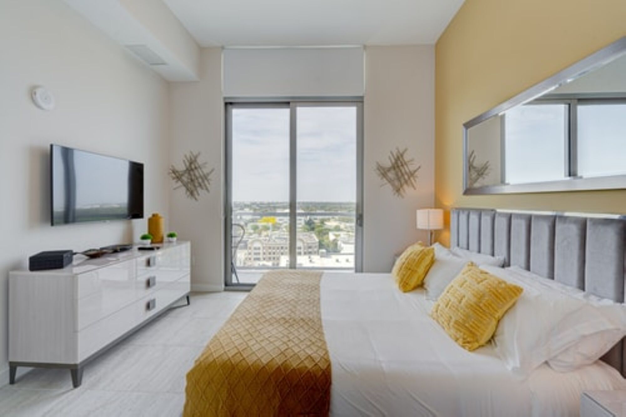 Quarto de hotel com cama, televisão, cômoda e uma grande janela com vista para a cidade.