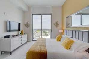 Quarto de hotel com cama, televisão, cômoda e uma grande janela com vista para a cidade.