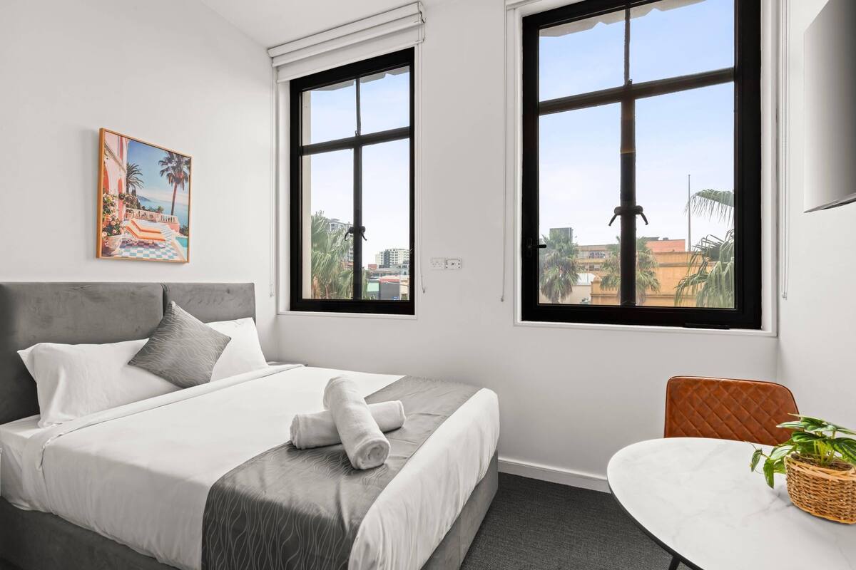 Quarto de hotel com cama, mesa de jantar e janelas amplas com vista para palmeiras e edifícios.