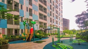 Um parquinho com escorregadores e equipamentos de ginástica, cercado por um edifício com sacadas e um jardim.