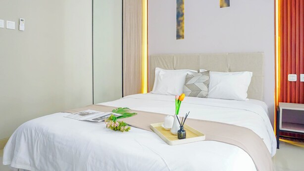 Cama bem arrumada com roupa de cama branca, mesa de cabeceira com abajur e uma obra de arte na parede.
