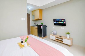 Quarto de hotel com cama, televisão, pequena cozinha e mesa de cabeceira com um vaso.
