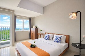 Quarto de hotel com cama, mesa de cabeceira, abajur, vista para o verde lá fora e janela com cortinas.