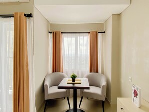 Quarto com duas cadeiras, uma mesa com um livro e uma planta, e uma janela com cortinas.