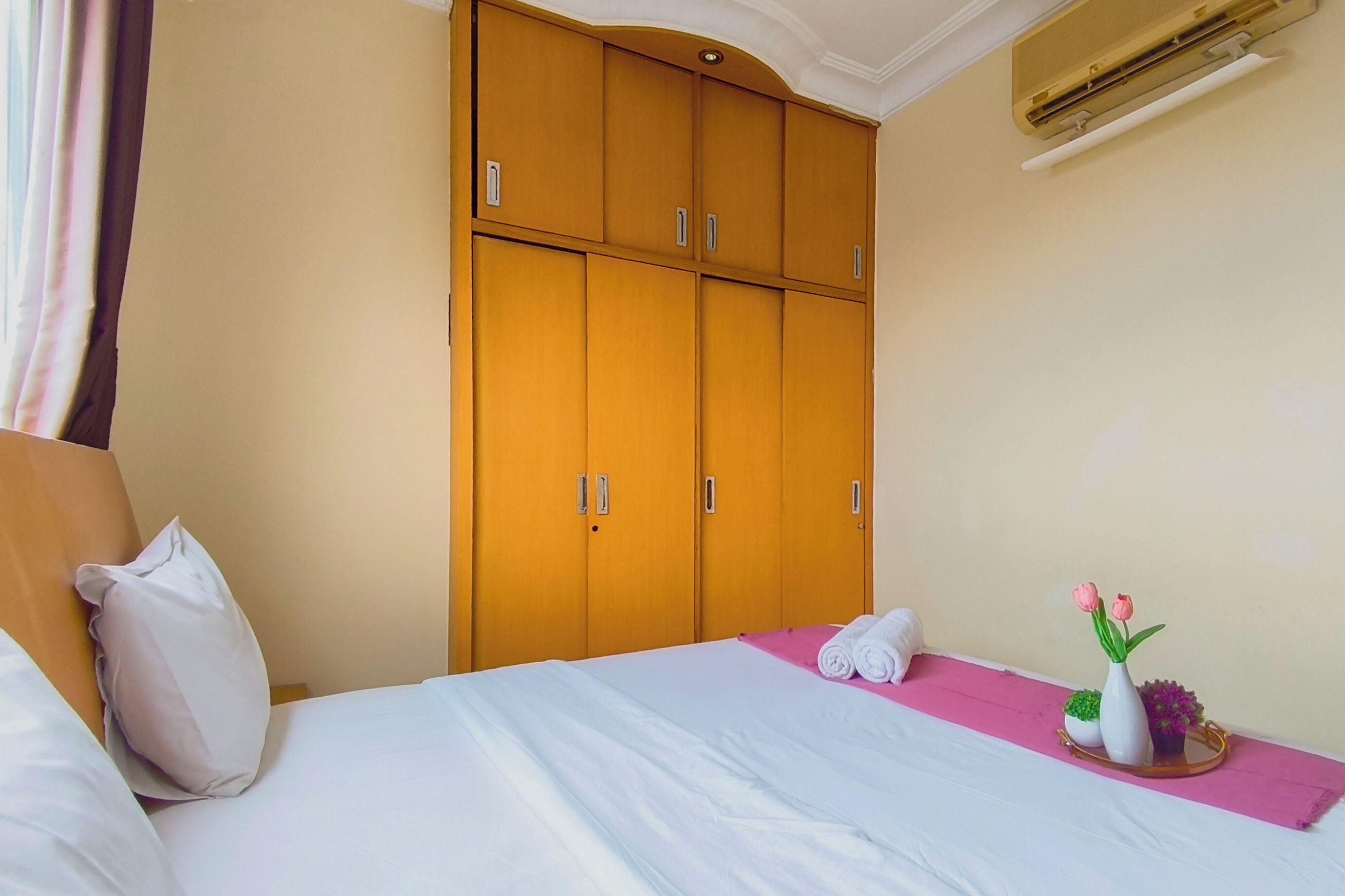 Quarto de hotel com cama, guarda-roupa, mesa de cabeceira com um vaso e flores, e uma toalha rosa.