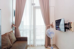 Quarto com sofá, ventilador de chão, televisão fixada na parede e cortinas translúcidas.