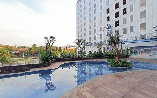 Piscina com deck de madeira, rodeada por plantas tropicais, e um grande edifício de hotel ao fundo.