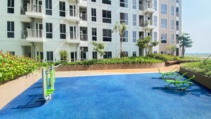 Piscina no terraço com piso azul, cercada por um muro baixo com vegetação e um edifício com varandas ao fundo.