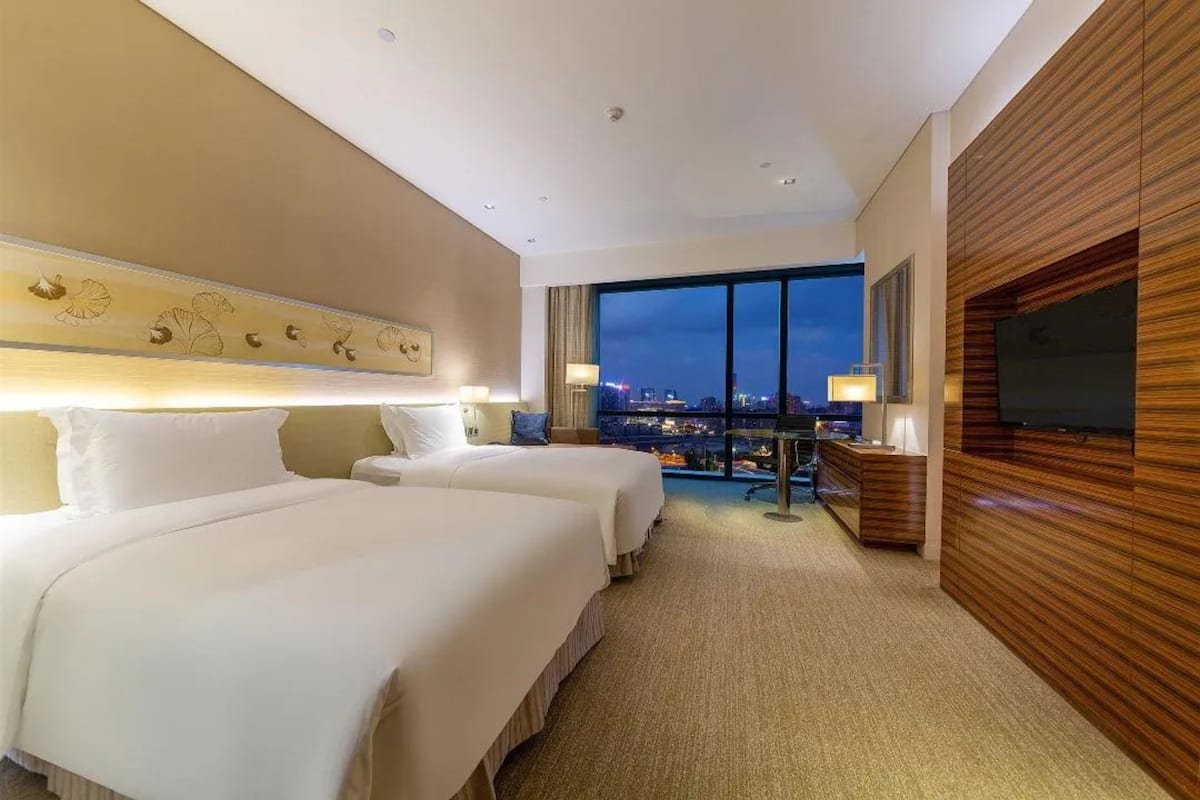 Quarto de hotel com duas camas, uma janela grande com vista para a cidade, uma televisão de tela plana e um painel de parede em madeira.