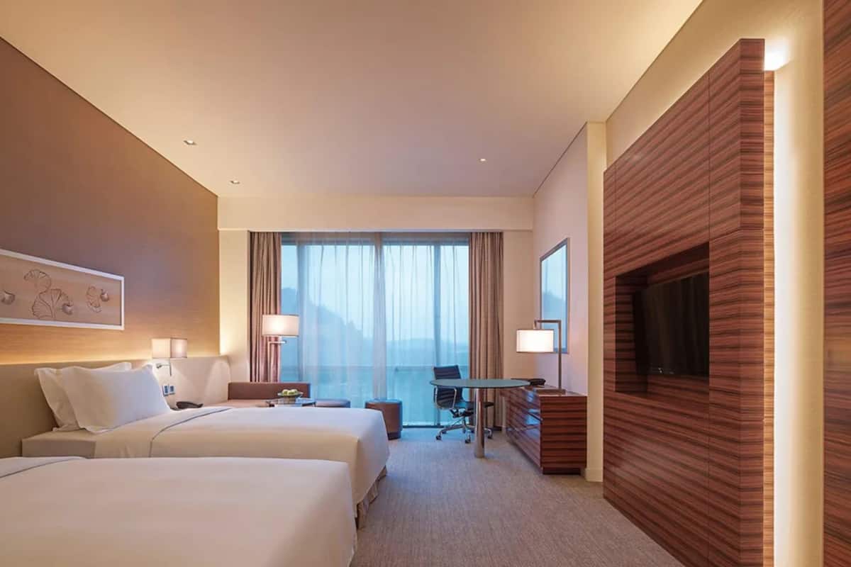 Quarto de hotel com duas camas, uma escrivaninha, uma cadeira, uma televisão e uma janela grande.
