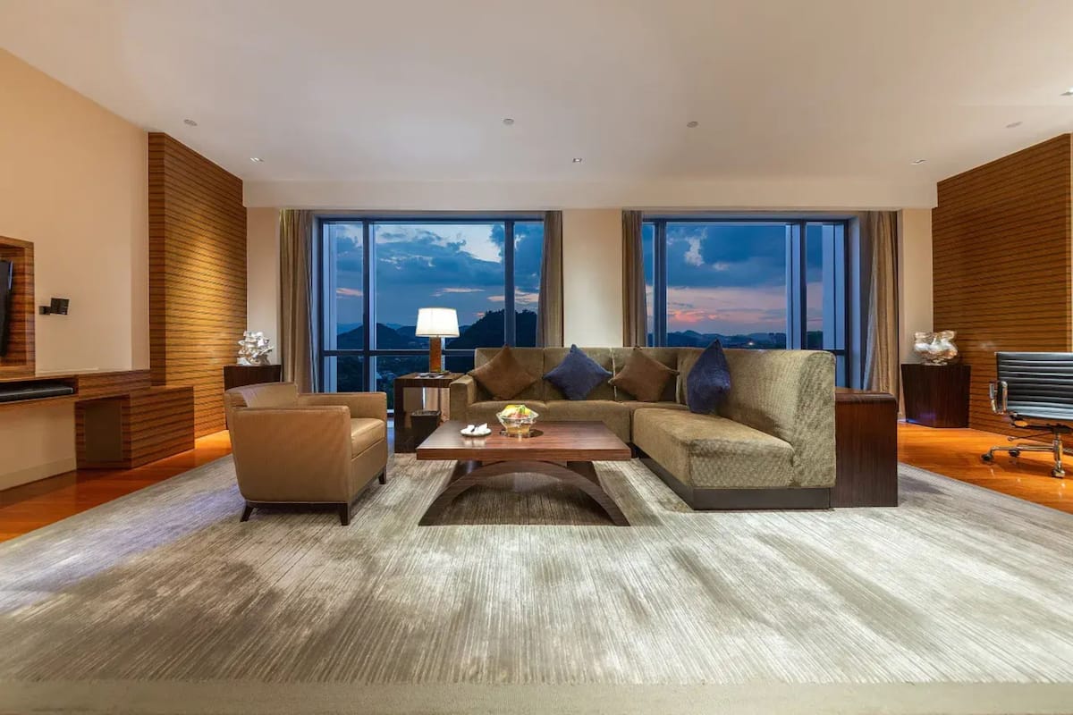Uma sala de estar moderna com sofá, poltrona, mesa de centro e televisão. Grandes janelas proporcionam vista do céu e das montanhas.