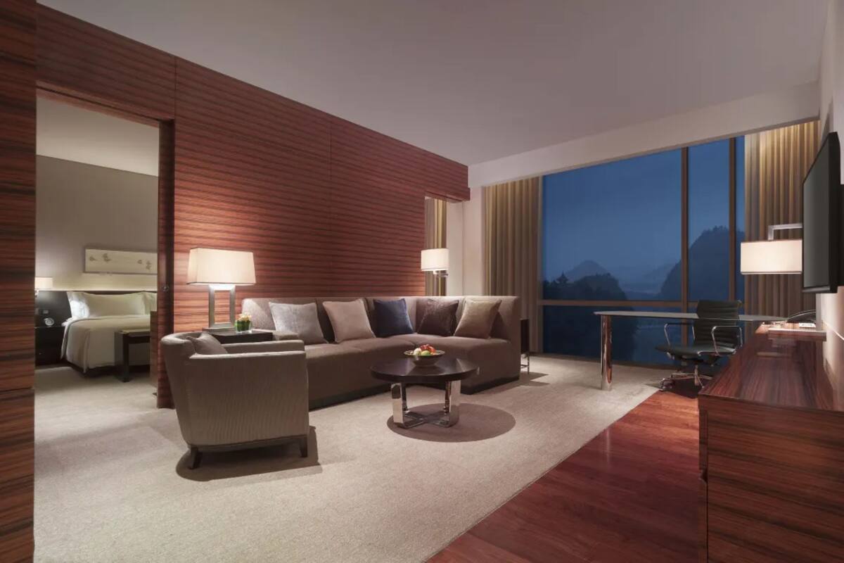 Quarto de hotel moderno com uma janela grande, um sofá, uma poltrona, uma mesinha e uma televisão.