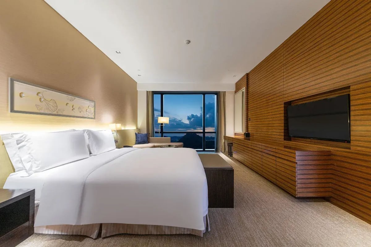 Quarto de hotel moderno com uma cama grande, uma TV de tela plana na parede, uma janela ampla com vista e uma parede de destaque em madeira.