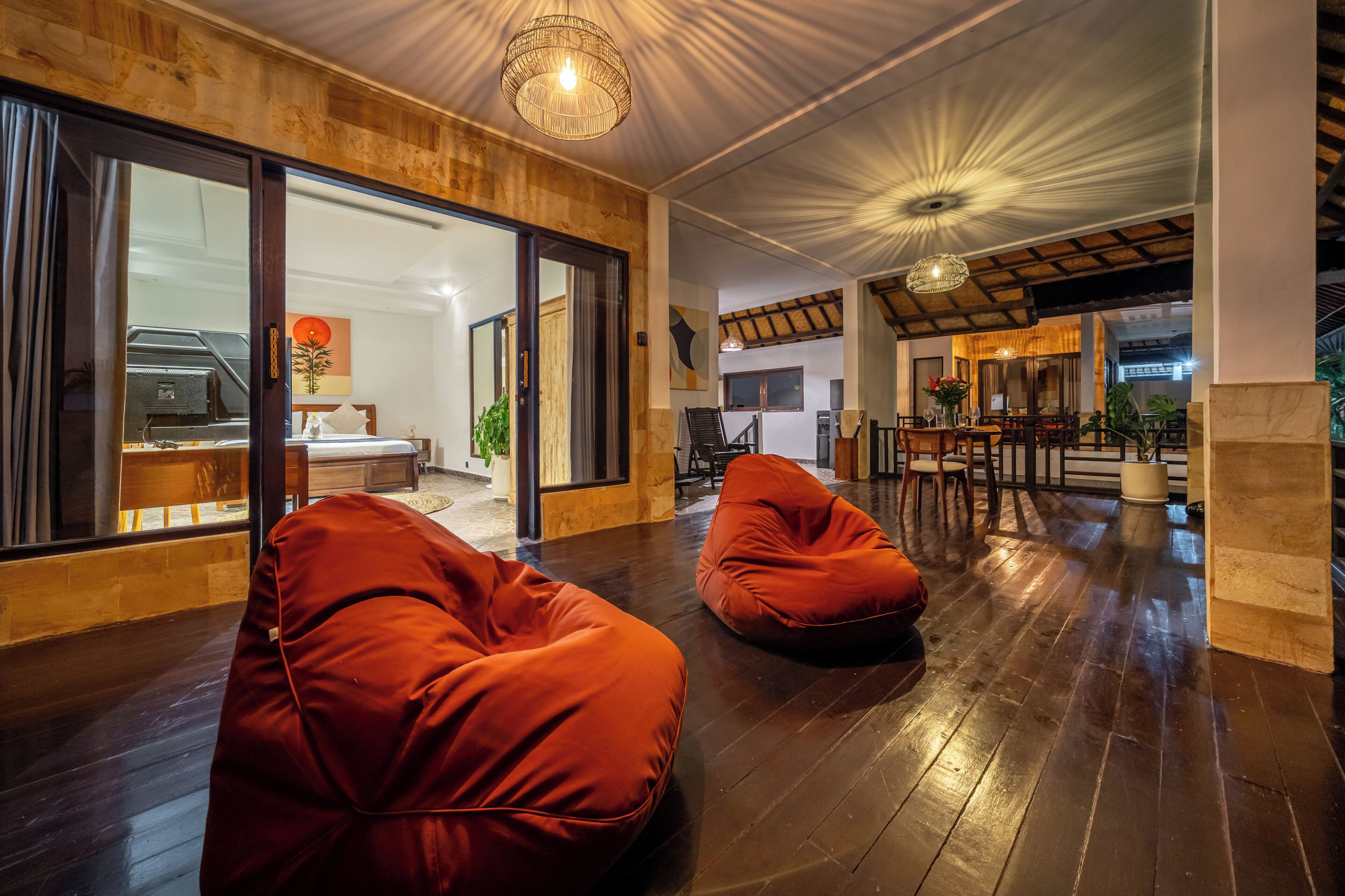 Um quarto espaçoso com uma grande porta de vidro deslizante que dá para uma varanda, uma área de jantar com mesa e cadeiras, e dois pufes.