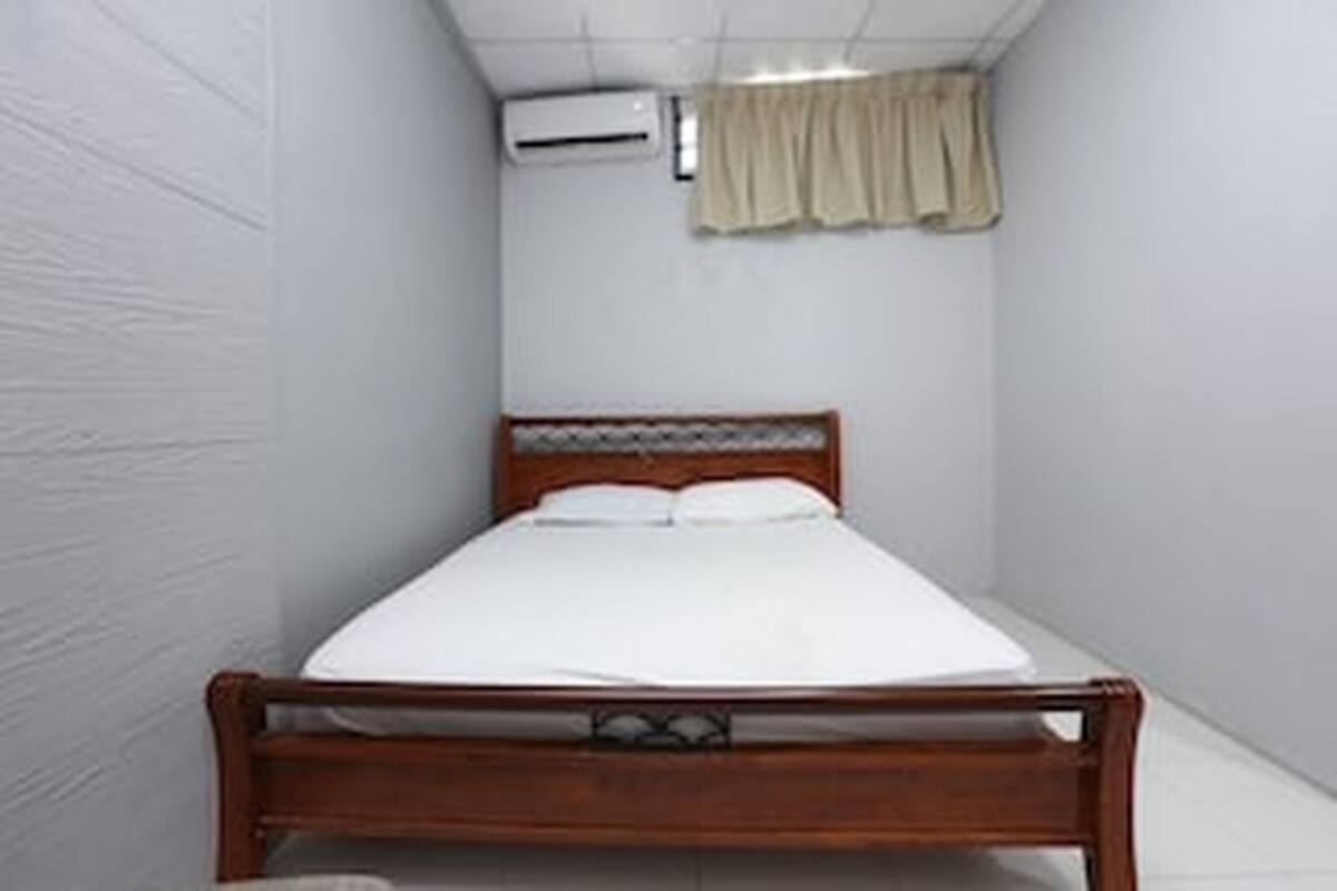 Uma cama de solteiro com estrutura de madeira e roupa de cama branca num quarto pequeno com paredes brancas e uma janela com cortina.