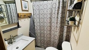 Casa de banho com sanita, lavatório, espelho, cortina de chuveiro e uma pequena prateleira com artigos de higiene pessoal.