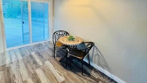 Quarto com piso de madeira, uma mesa redonda com cadeiras, luminária no teto e uma porta de vidro deslizante que dá para o exterior.