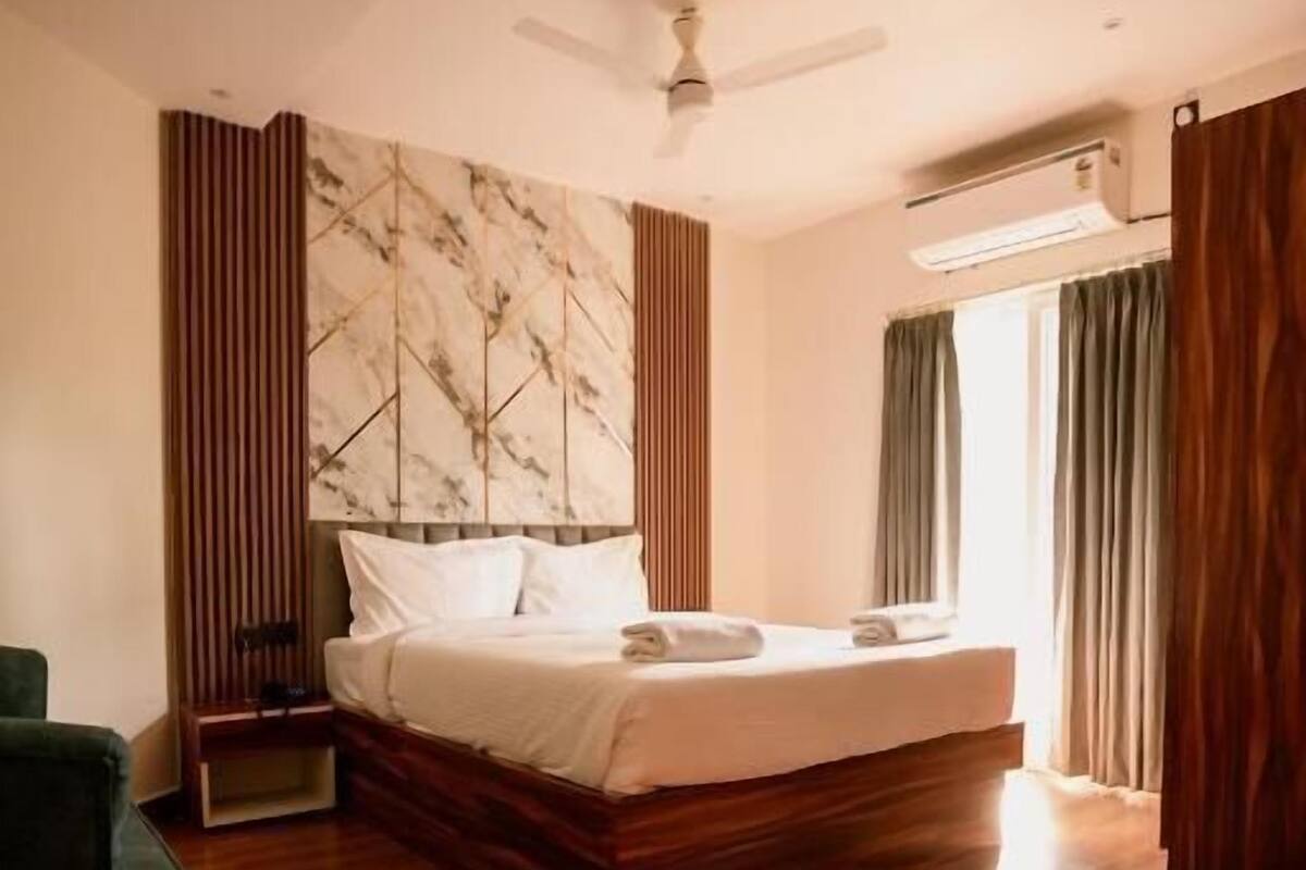Quarto com cama, ventoinha no teto, mesa de cabeceira, poltrona verde e piso de madeira.